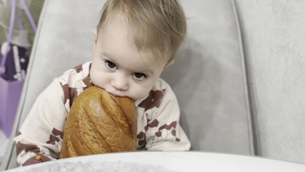 Hungry baby sits on a chair and munches on an oversized loaf of bread