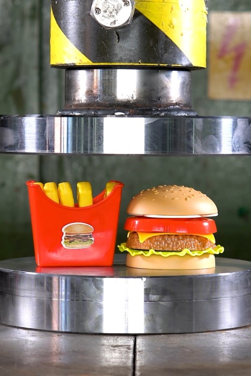 Una prensa hidráulica aplasta una hamburguesa de juguete con patatas fritas