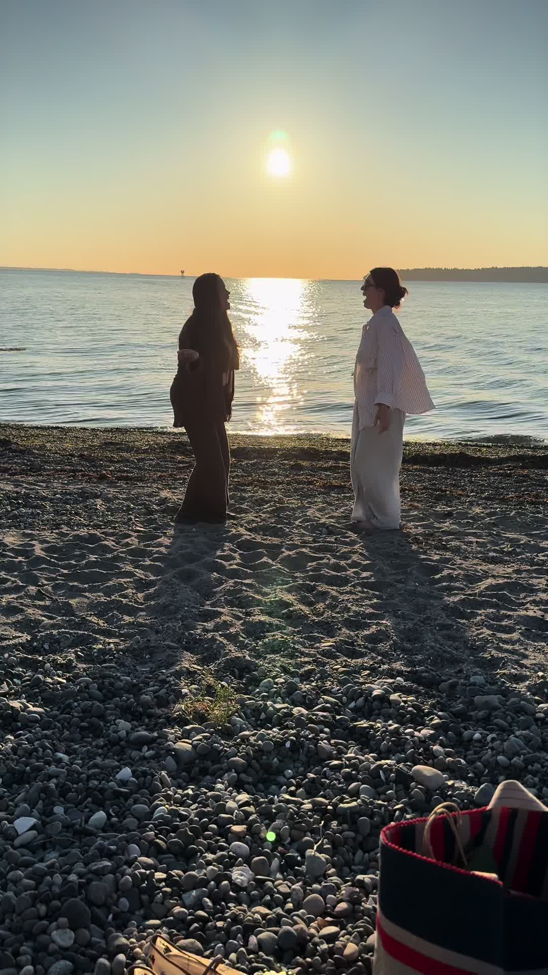 Two friends dancing joyfully on a pebbled beach as the sun sets over ...